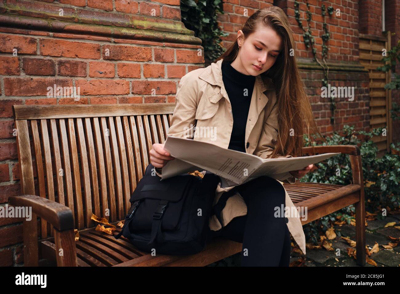 Attractive girl reading newspaper sitting with backpack on bench ...