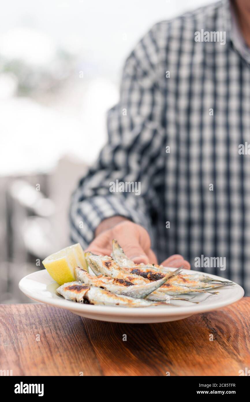 Waiter in a plaid shirt serving a plate of fish on the beach. Wooden ...
