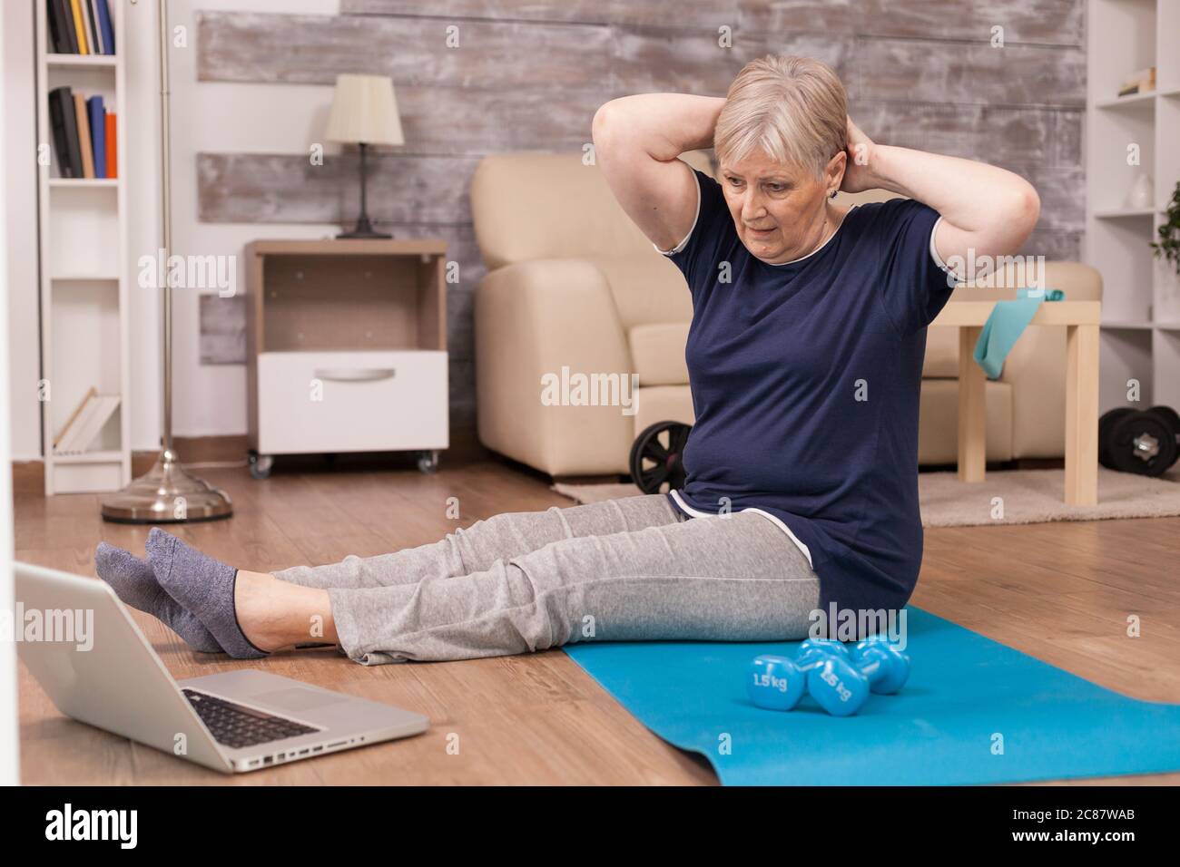 Aged woman doing online training sitting on yoga mat at home. Old person pensioner online ...