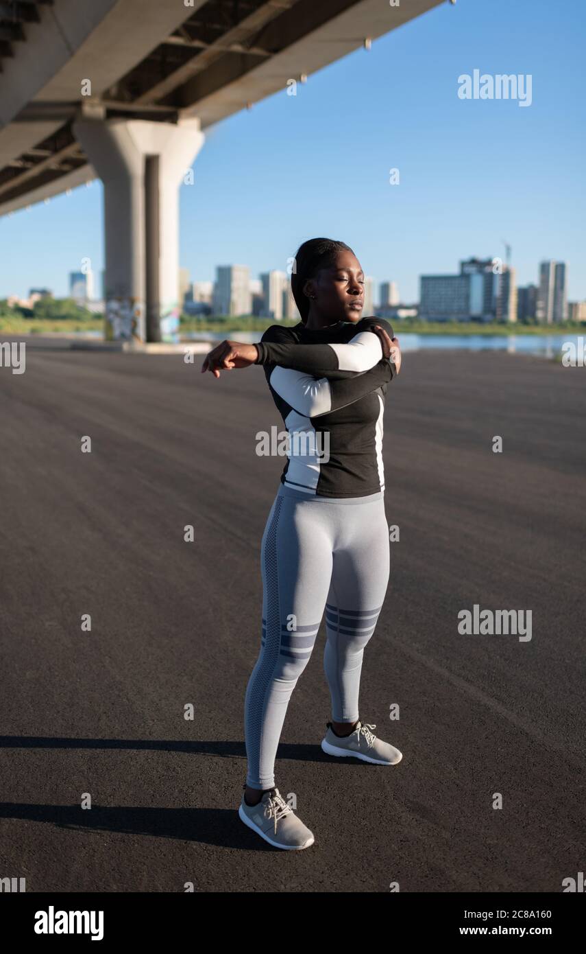 Black female runner stretching arms during morning workout under bridge ...