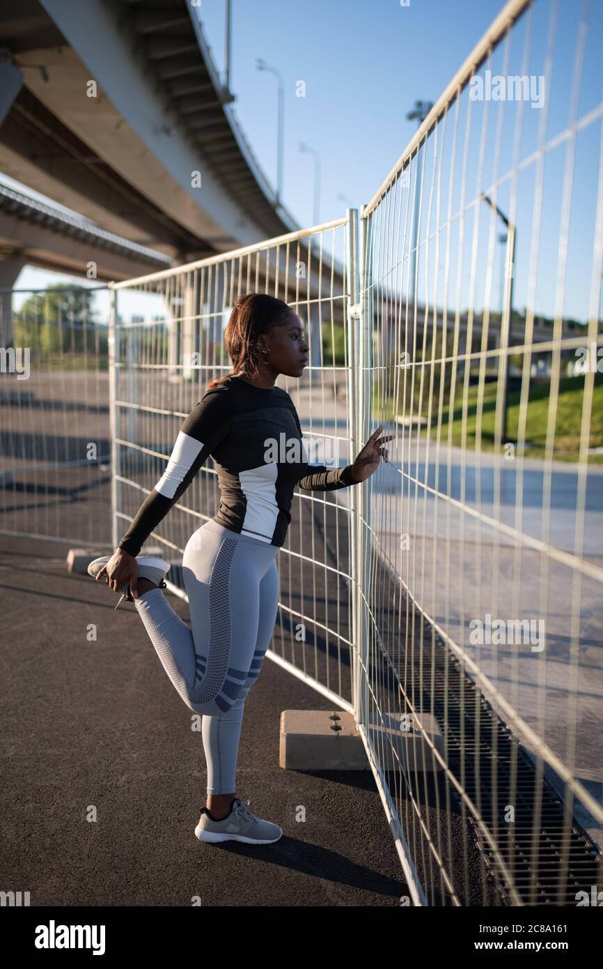 Side view of ethnic runner stretching leg near fence during morning ...