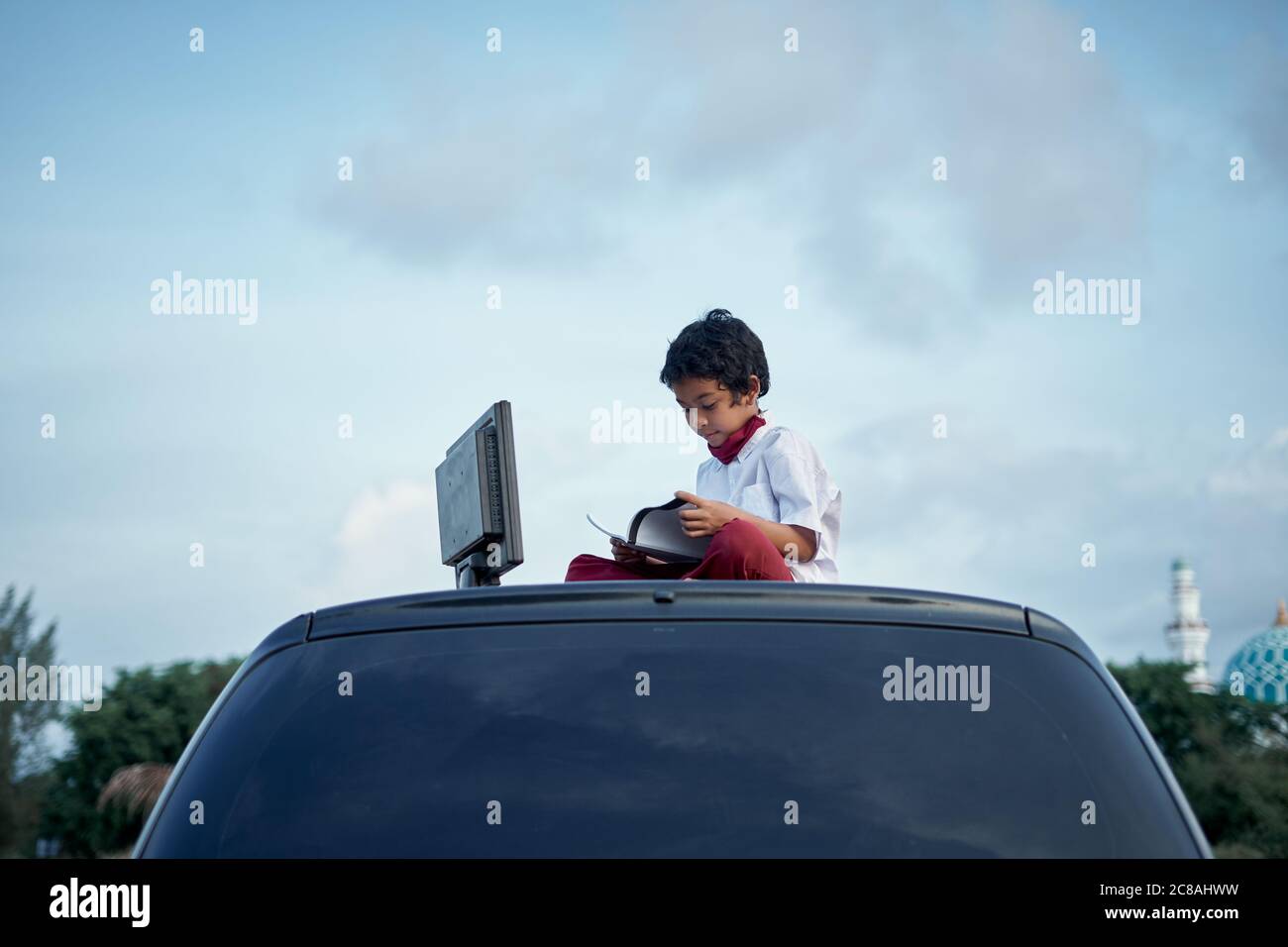 Schoolboy doing homework using computer on top of car roof, New Normal ...
