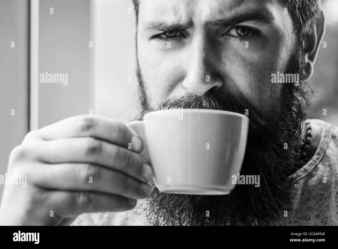Grumpy looking bearded man drinking morning cup of coffee near window ...