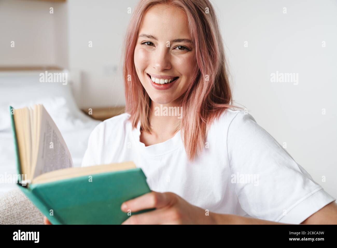 Image of joyful pretty woman with pink hair reading book and smiling ...