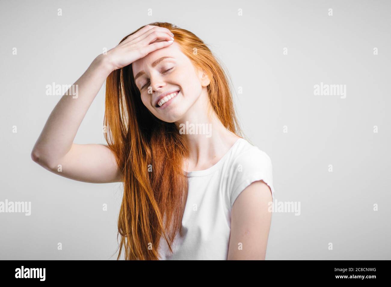 Happy girl with freckles smiling with closed eyes touching her red hair over white background ...
