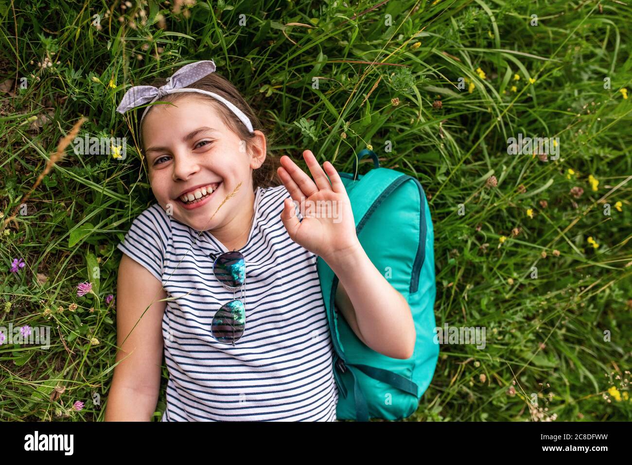Cute little girl smiling and waving hello while lying with her backpack ...
