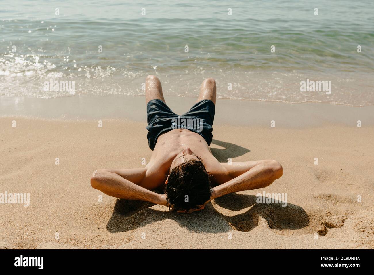 Shirtless young man with hands behind head lying on sand at beach ...