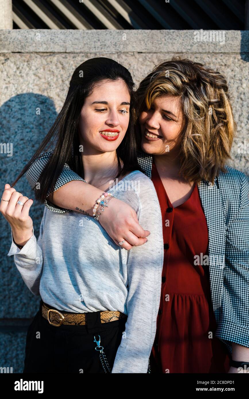 Smiling woman embracing girlfriend while standing against wall in city ...