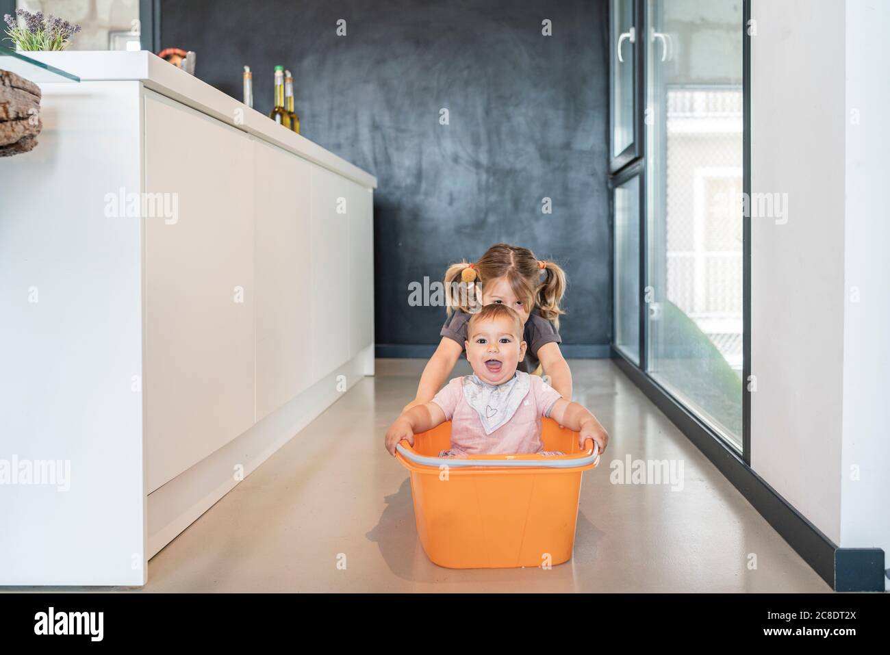 Girl pushing baby sister sitting in bucket on floor in modern kitchen ...