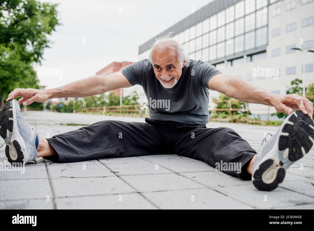 Smiling senior man stretching body while sitting on footpath in city ...