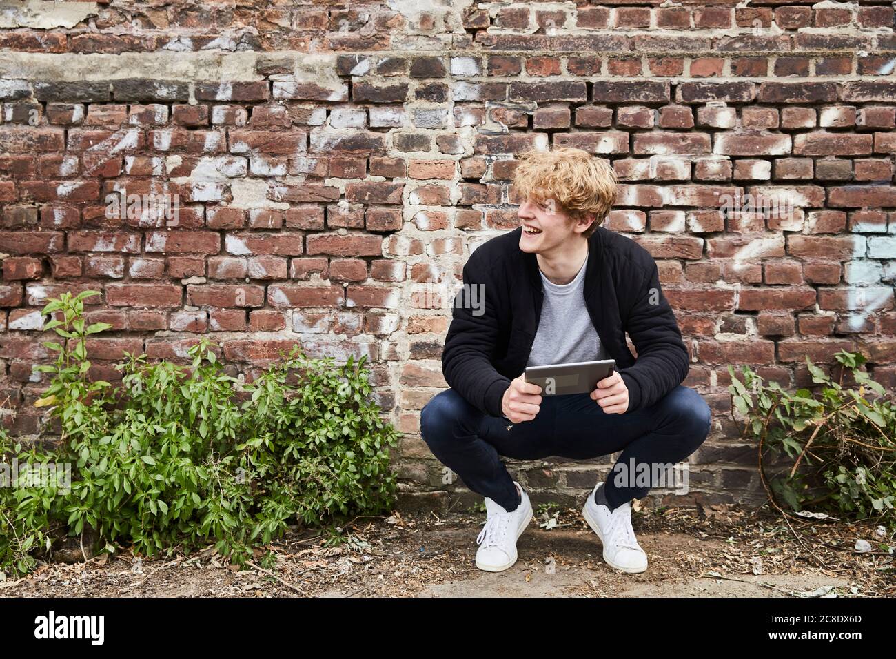 Laughing young man crouching in front of brick wall with digital tablet ...