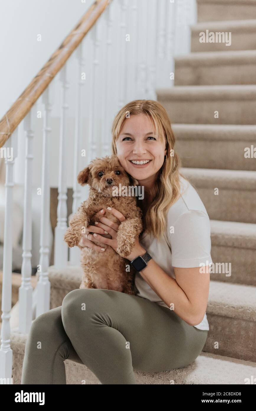 Smiling woman holding cute poodle while sitting on steps at home Stock ...