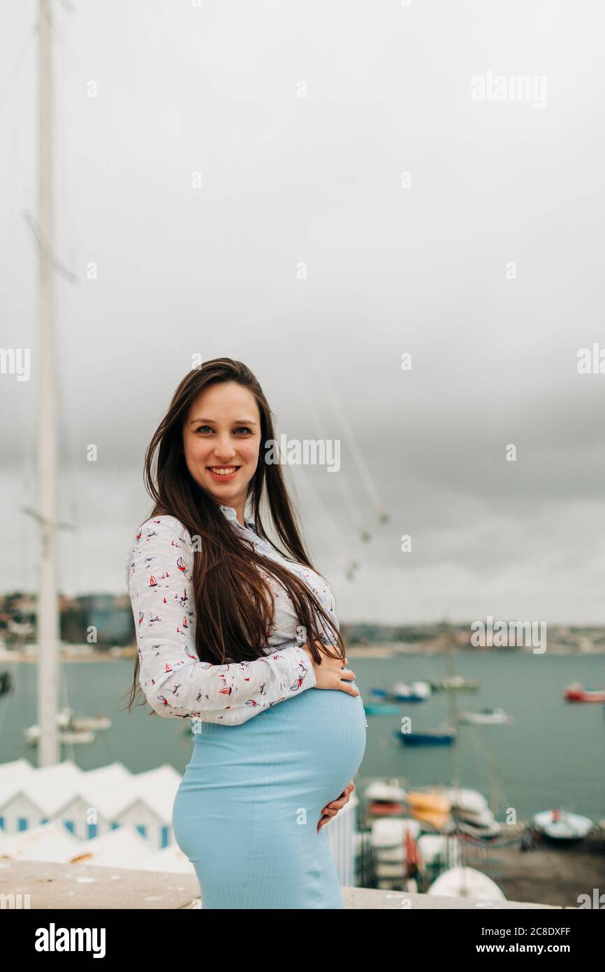 Happy pregnant woman holding belly while standing on pier at harbor ...