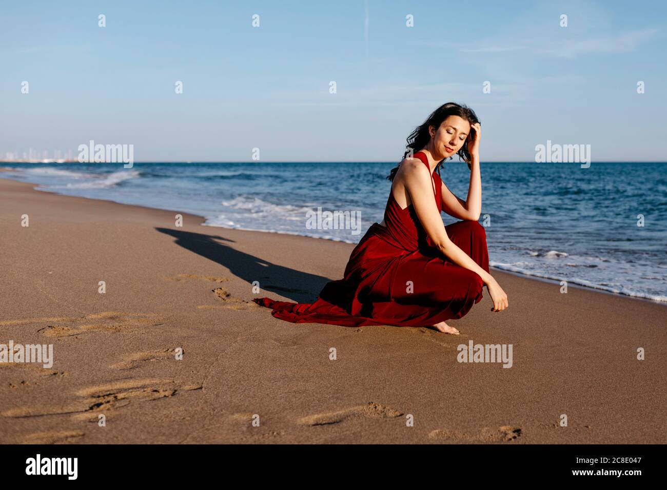 Delicate woman in red dress sittingh on the beach, feeling the sun ...