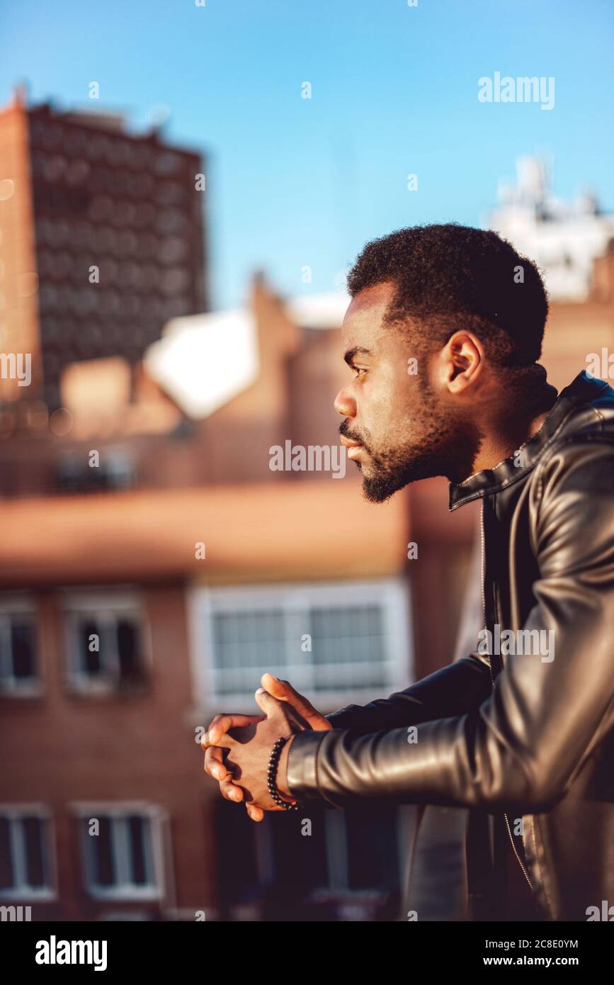 Thoughtful young man looking away while standing by railing on rooftop ...