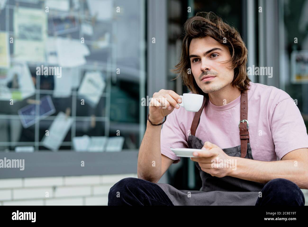 Thoughtful male barista holding coffee sitting outside coffee shop ...