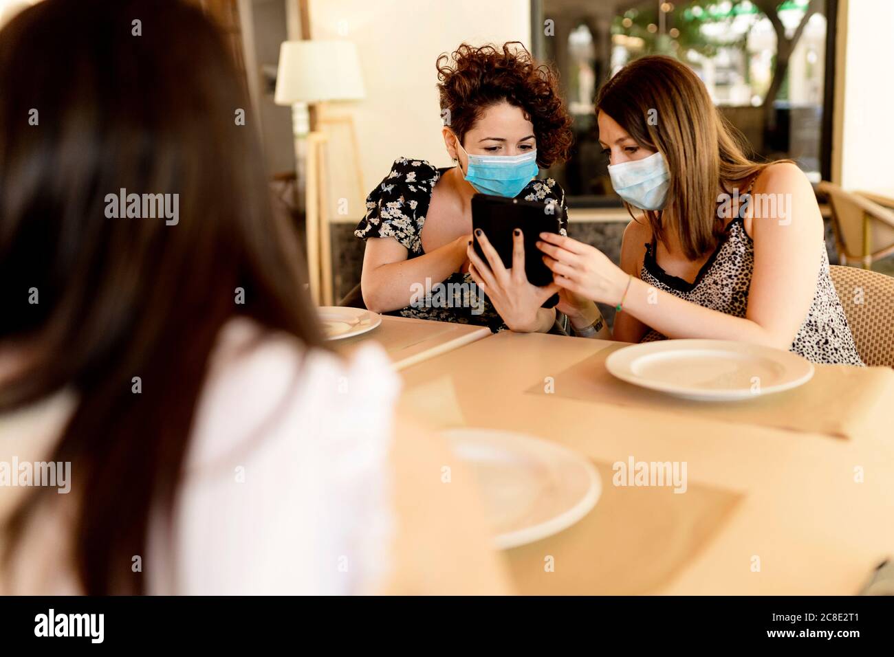 Women wearing masks while sharing digital tablet sitting at table in ...