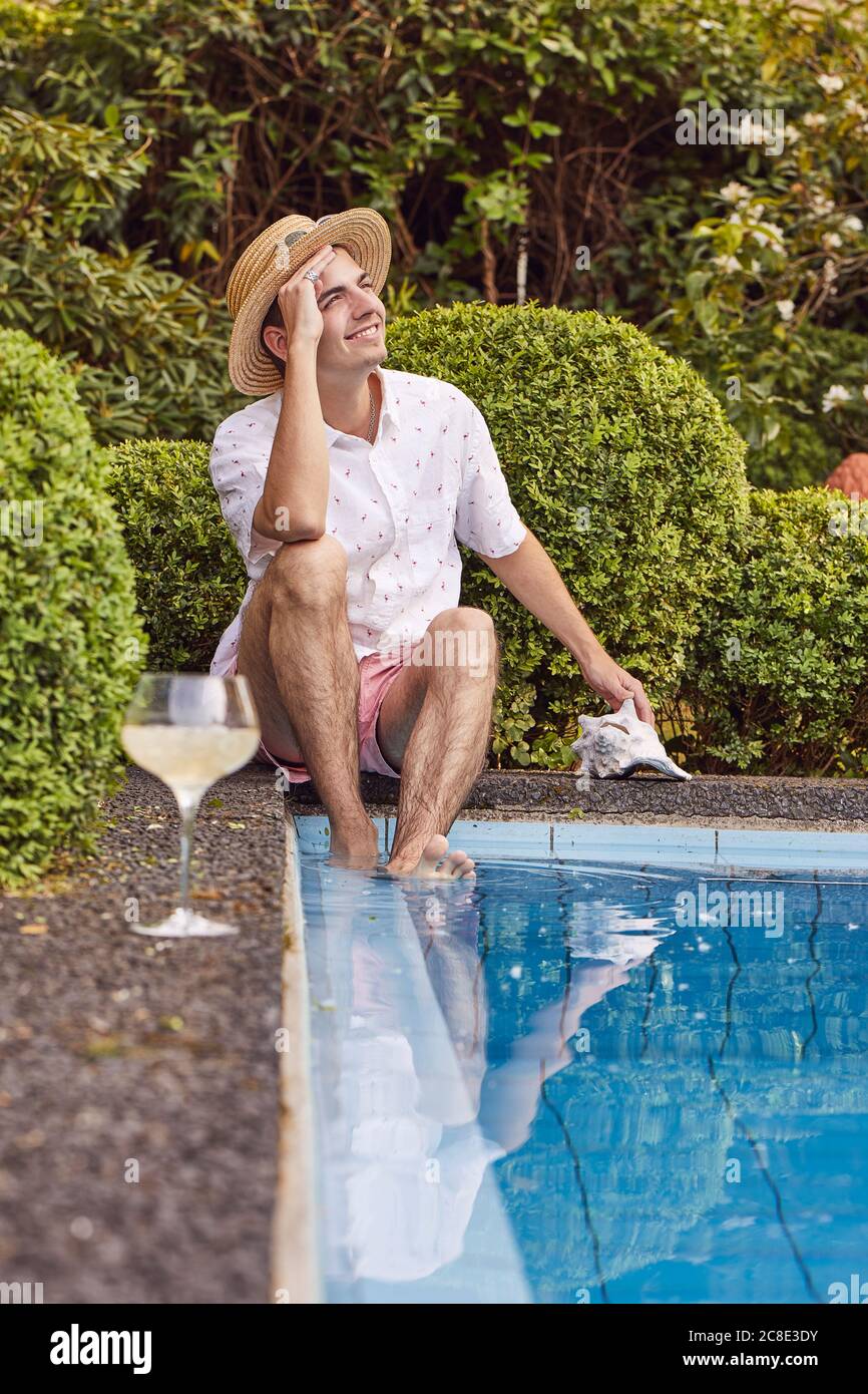 Thoughtful young man wearing hat sitting with conch shell at poolside ...
