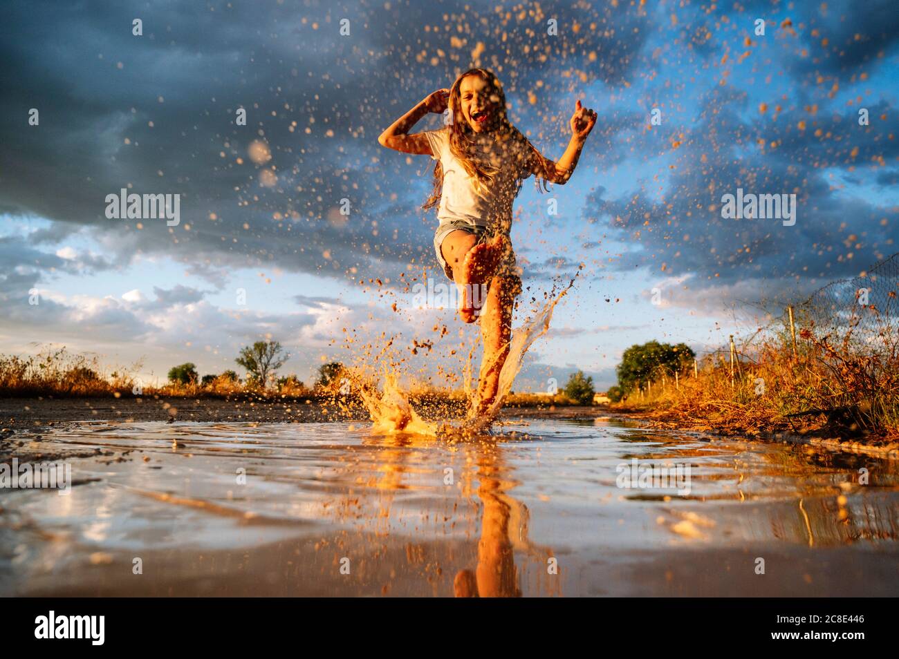 Happy girl playing while splashing water on puddle against cloudy sky ...