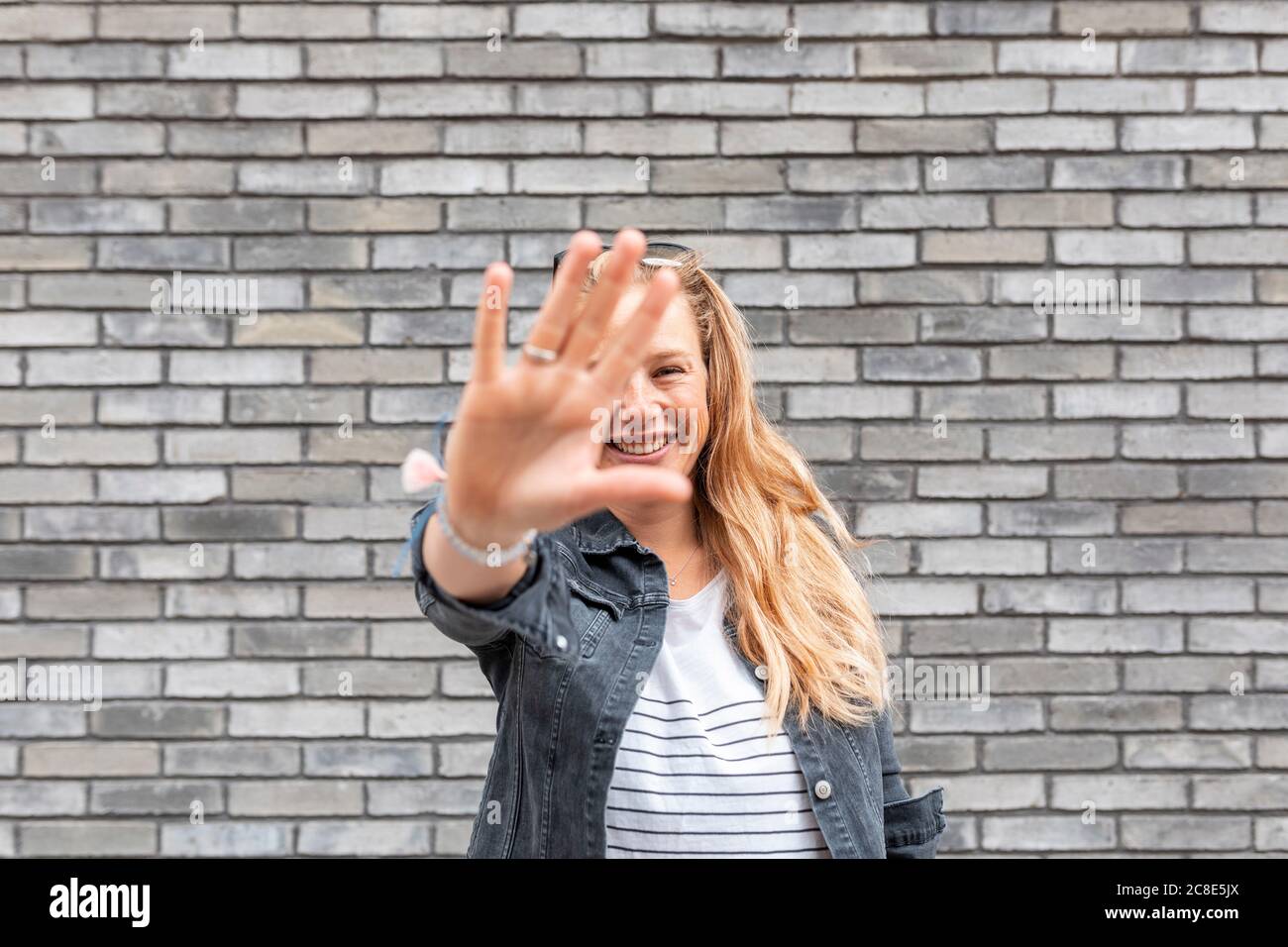 Smiling woman hiding face with hand while standing against gray brick ...