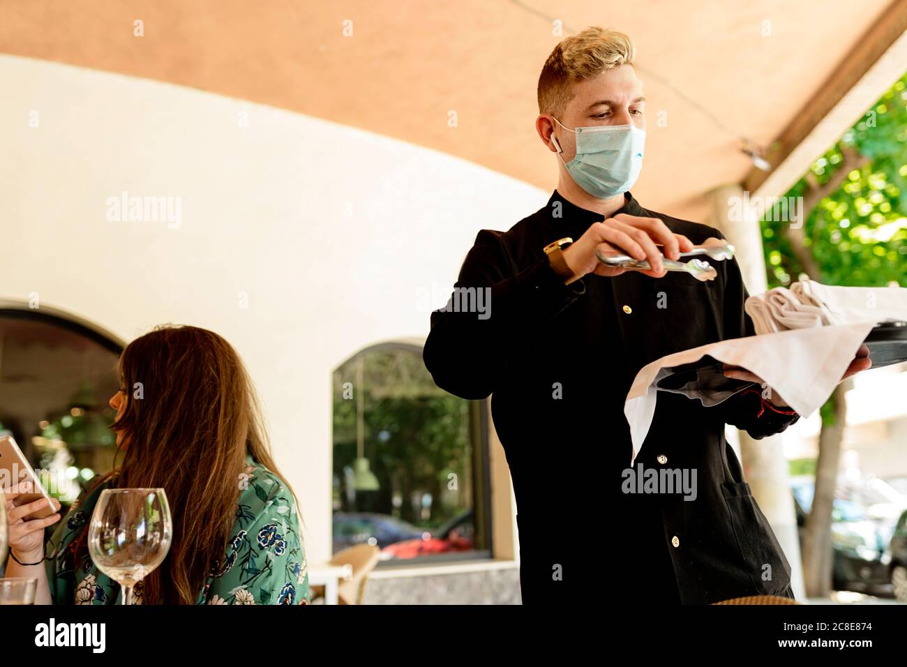 Young waiter holding serving tongs and napkins by female customer in ...