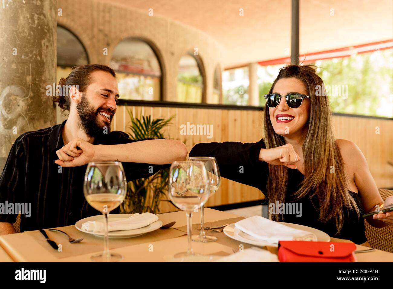 Happy young man and woman giving elbow bump while sitting at table in ...