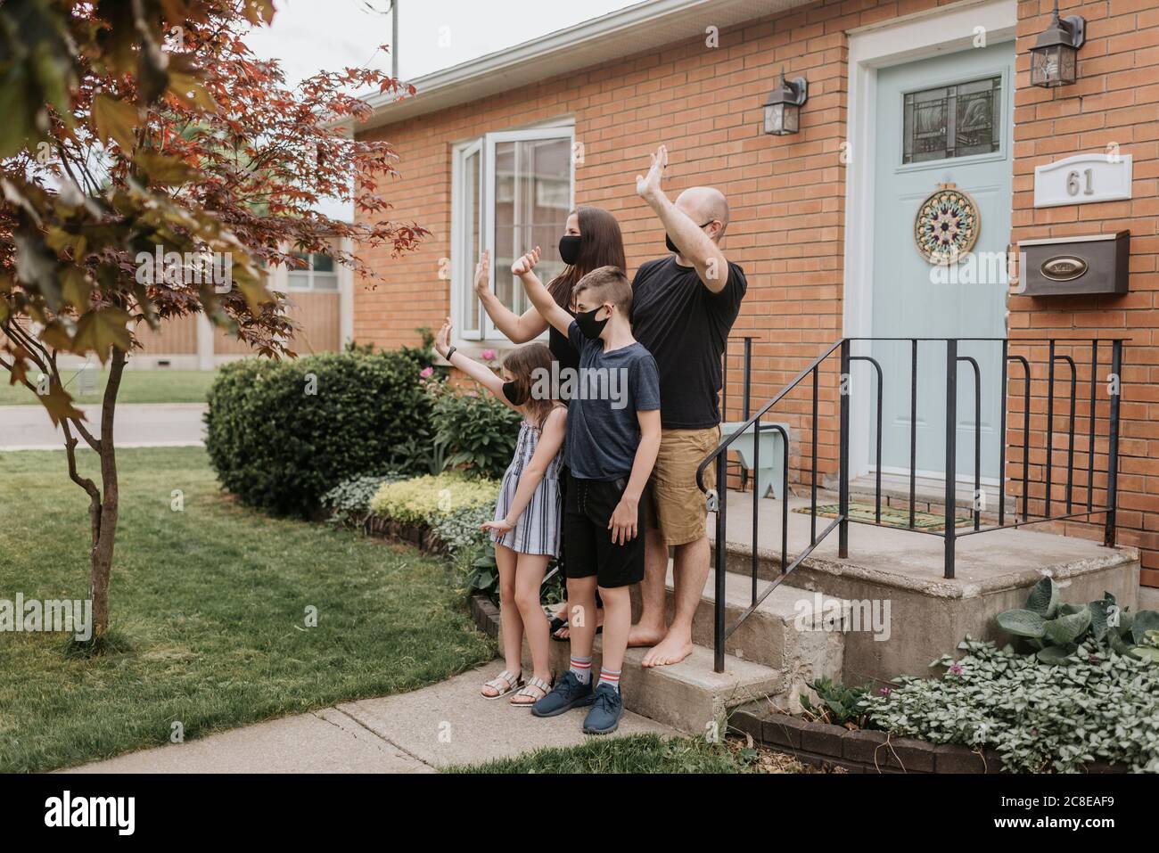 Family wearing masks waving hands while standing outside house in yard ...