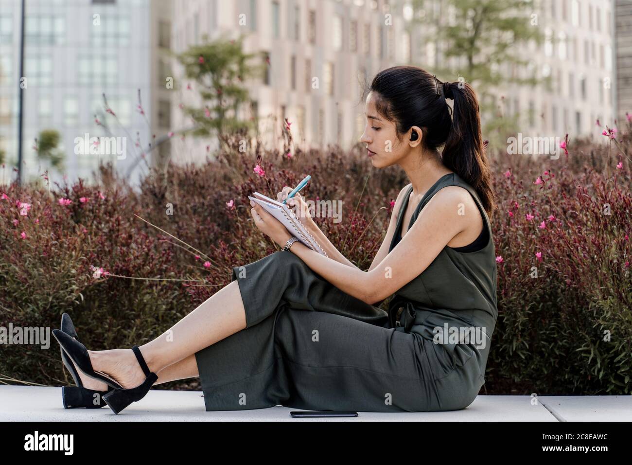 Female professional writing in note pad while sitting on seat by plants ...