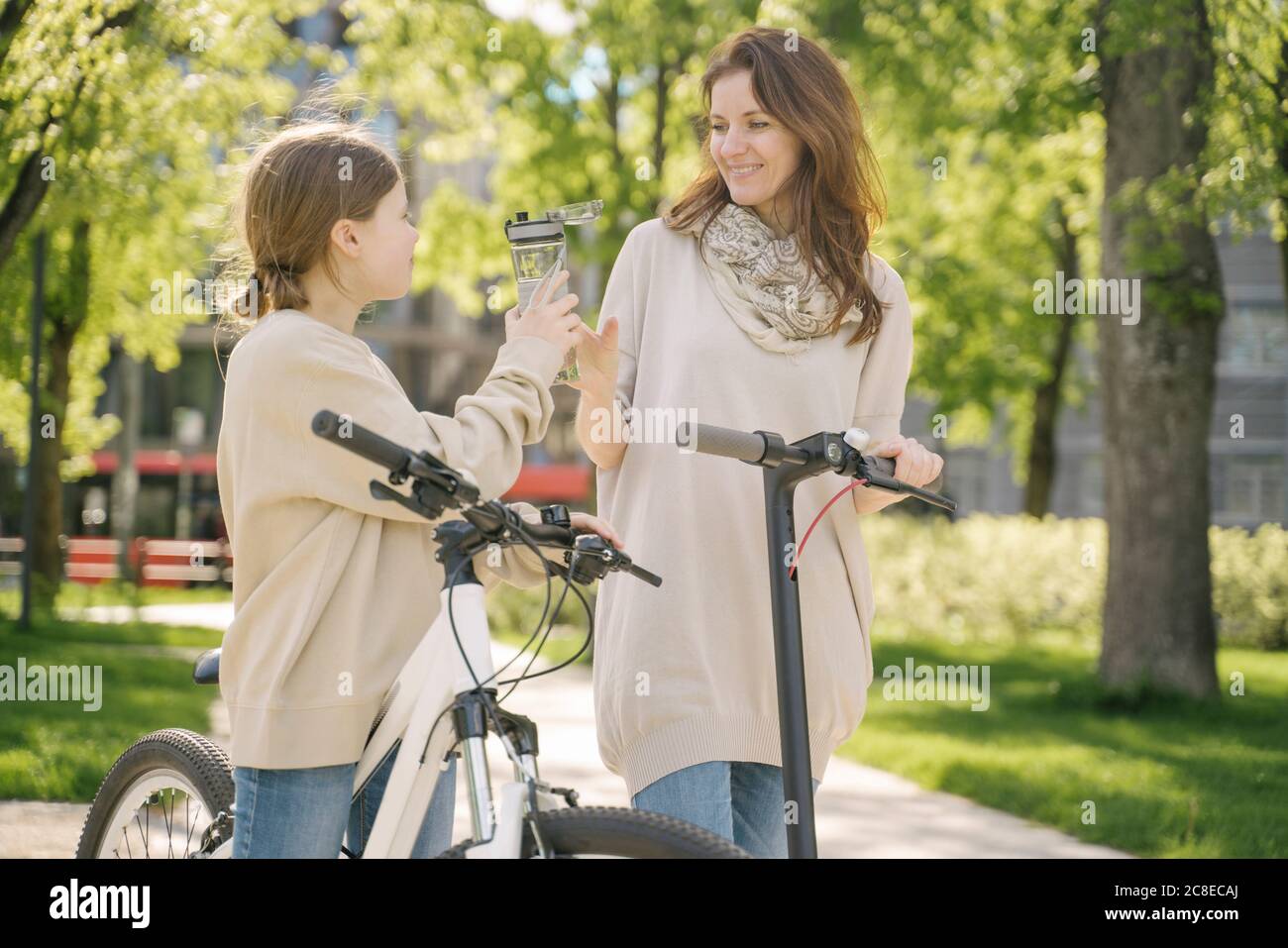 Girl sharing water with mother while standing in city park Stock Photo ...