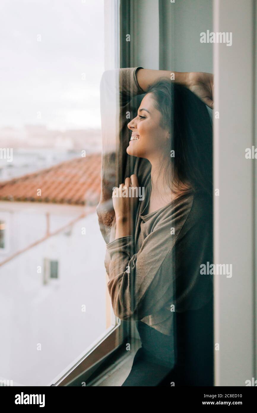 Smiling young woman looking through window at home seen through glass ...