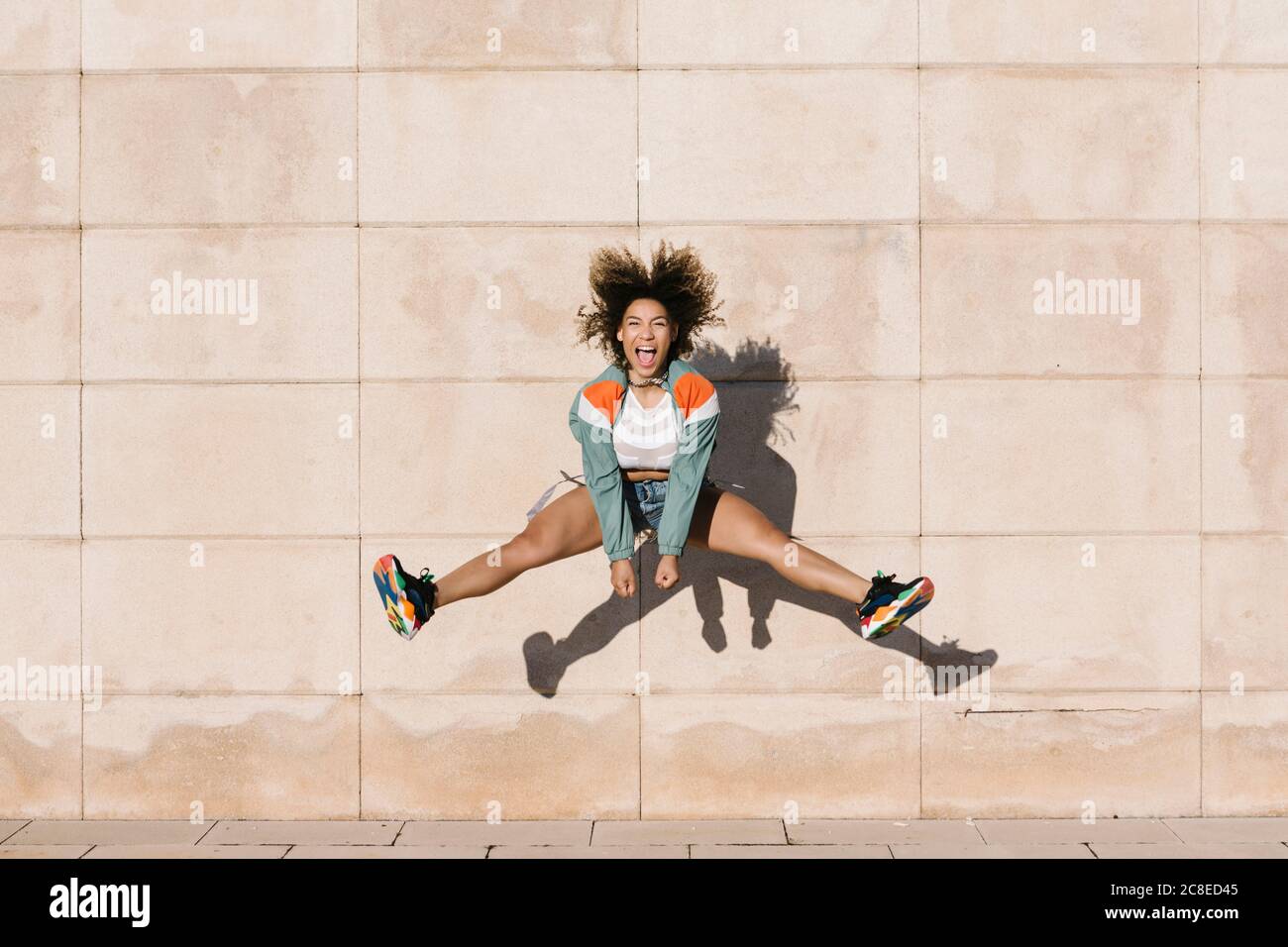 Excited young woman screaming while jumping against wall during sunny ...