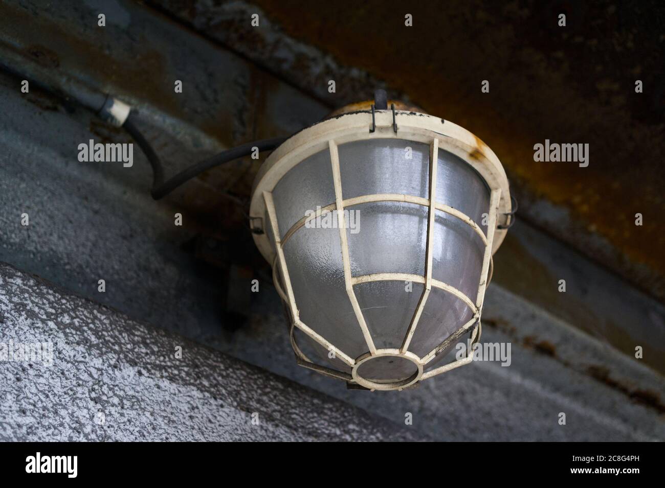 Industrial light is protected by white wire cage. Metal is rusted ...