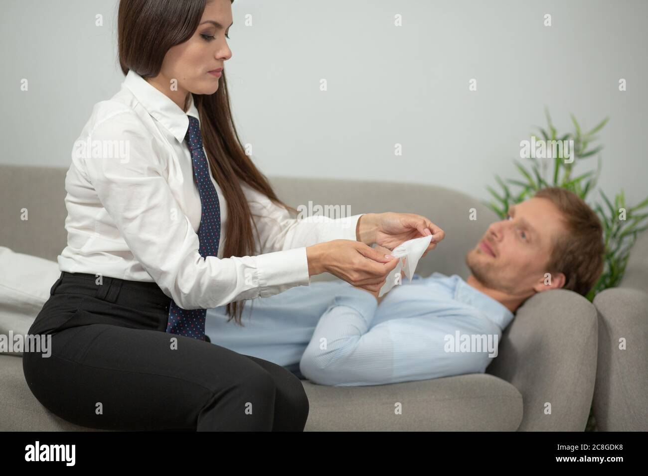 Woman putting mask on her husbands face. Young man laying on couch and ...