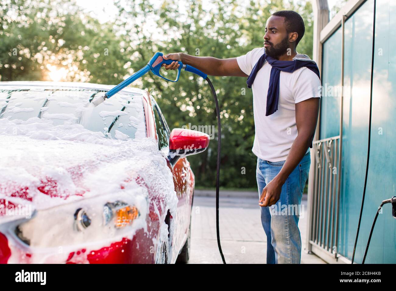 Portrait of handsome bearded young African man washing his red car with ...