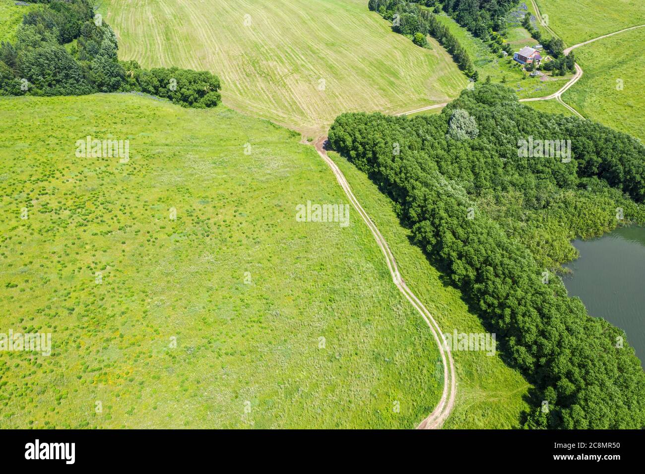 aerial view of a dirt road crossing through green fields near forest ...