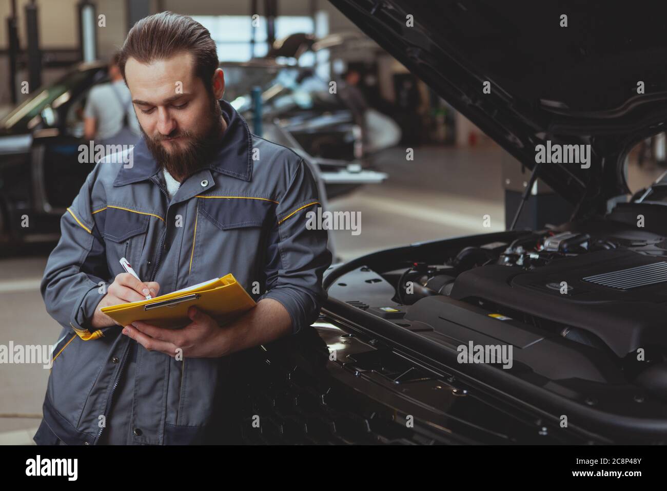Bearded male mechanic looking concentrated, writing on his clipboard ...