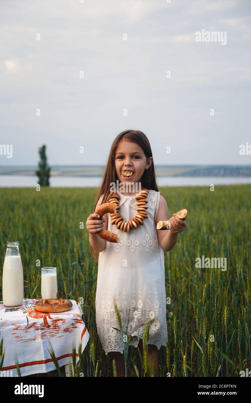 Cute smiling little girl eating bagel with milk in green field ...