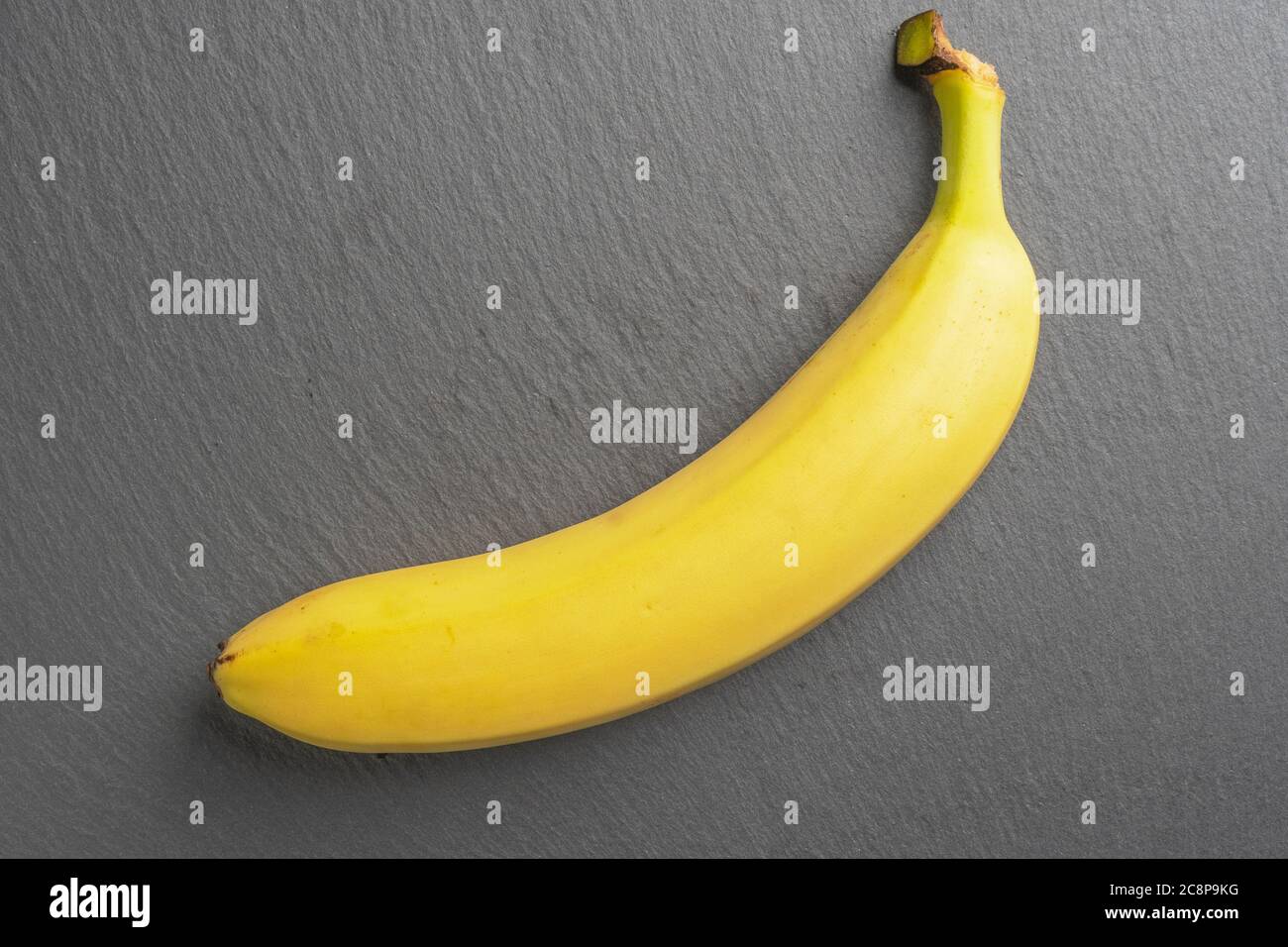 Ripe unpeeled banana with bright yellow peel on a stone surface. Gray ...