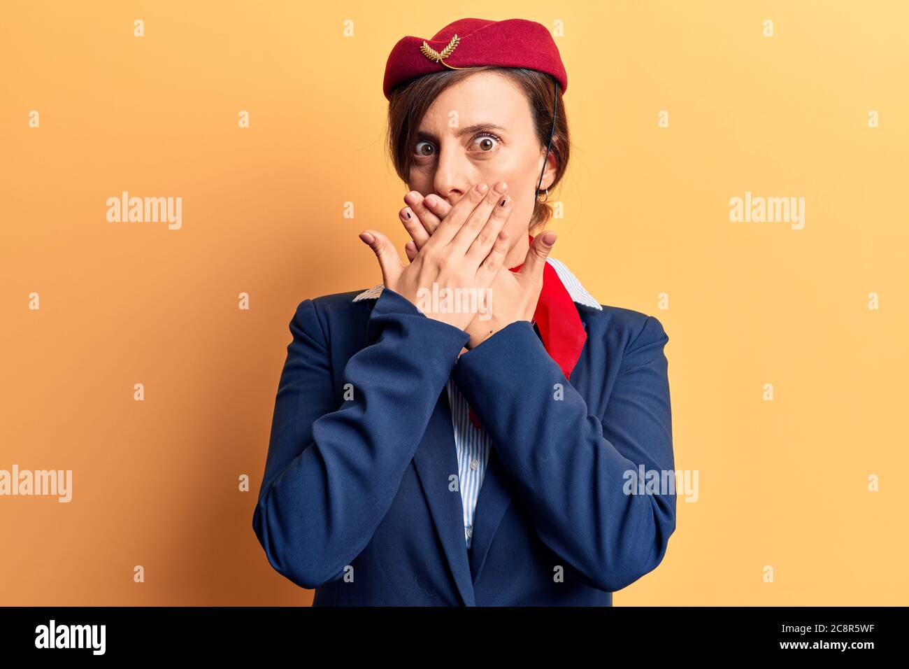 Young beautiful woman wearing stewardess uniform shocked covering mouth ...