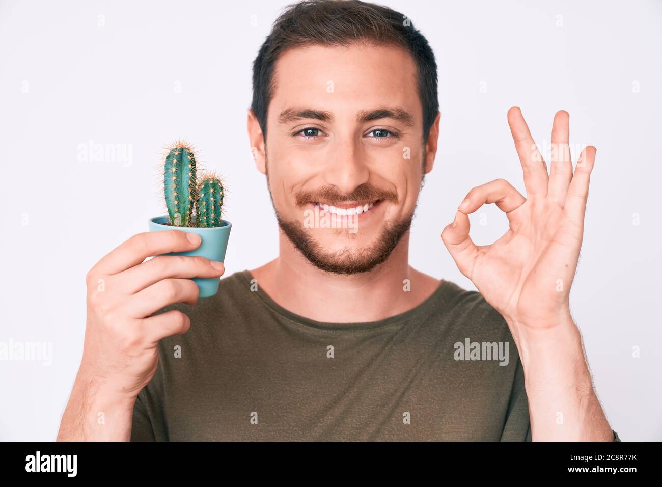 Young handsome man holding small cactus pot doing ok sign with fingers ...