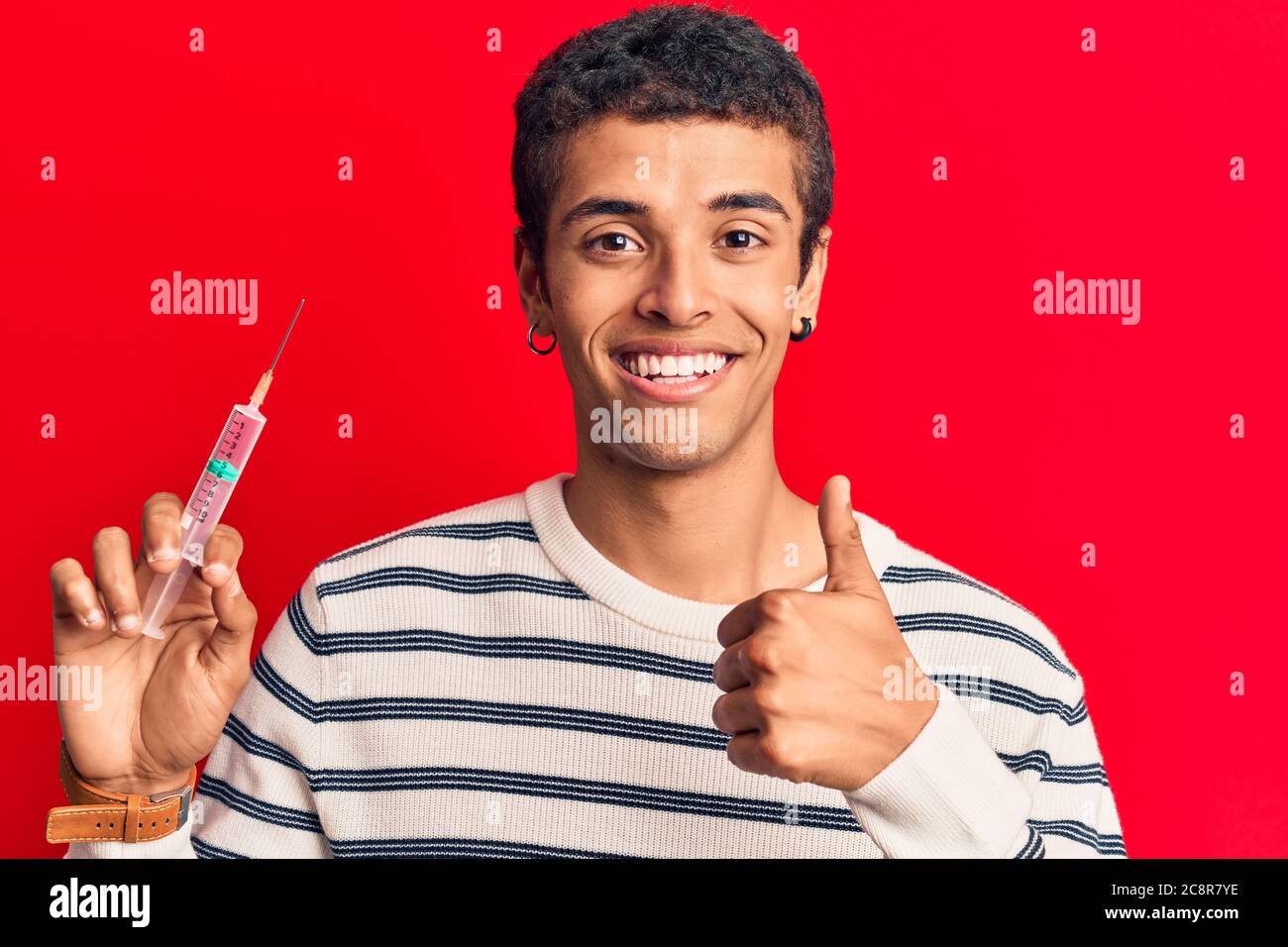 Young african amercian man holding syringe smiling happy and positive, thumb up doing excellent ...