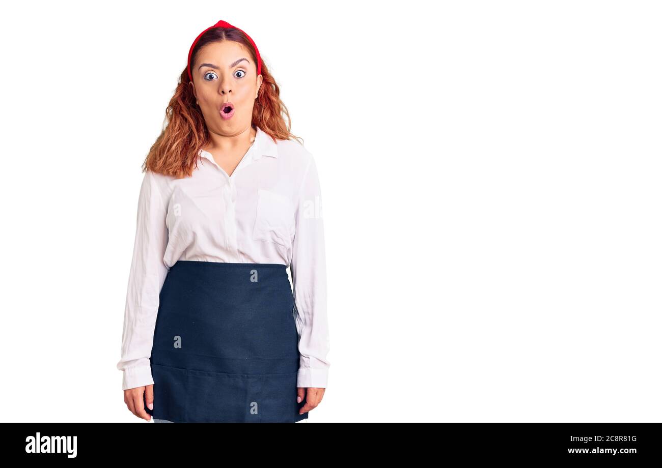 Young latin woman wearing waitress apron afraid and shocked with ...