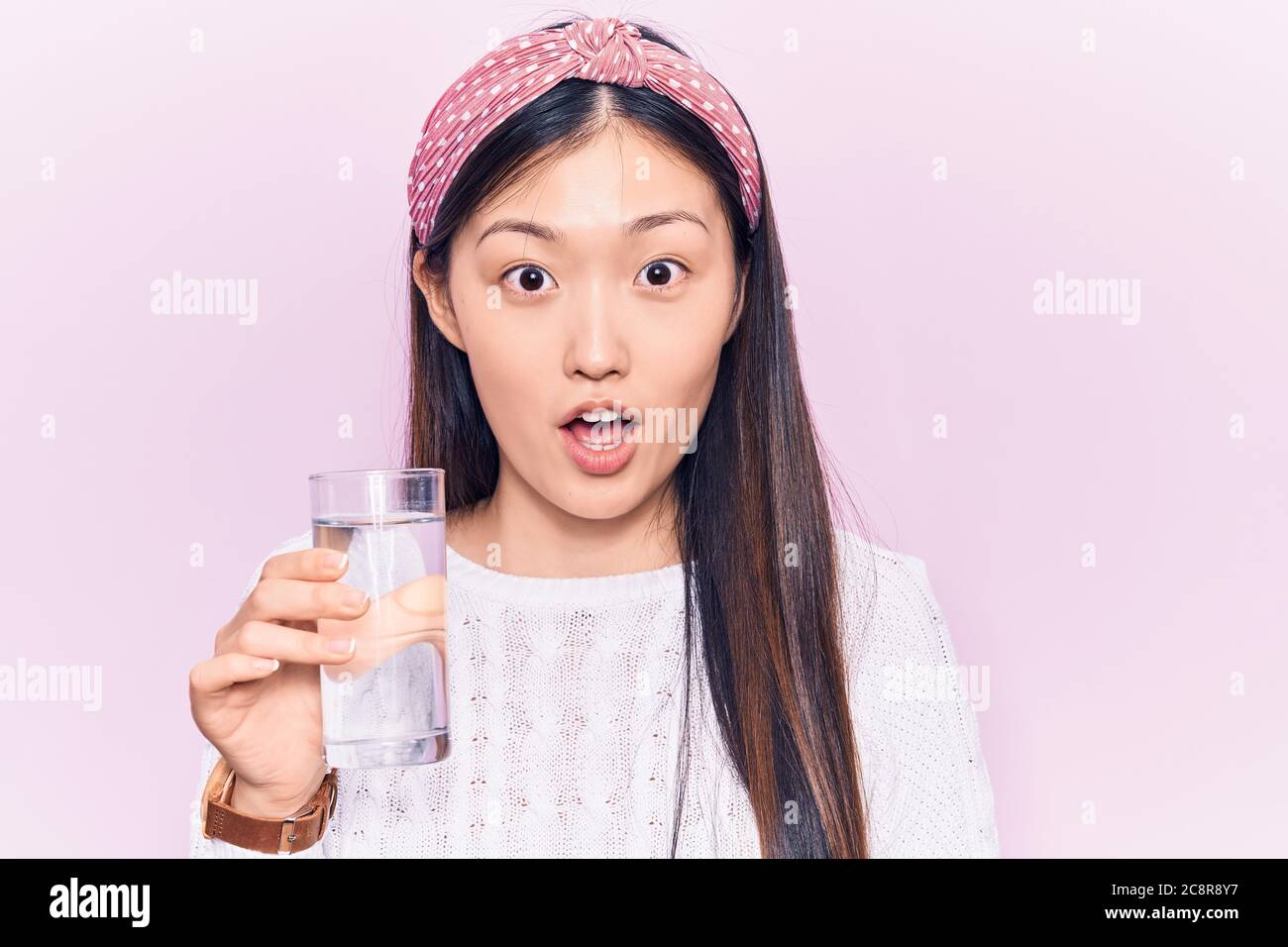 Young beautiful chinese woman drinking glass of water scared and amazed ...
