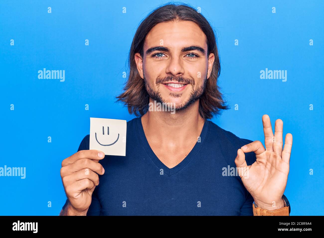Young handsome man holding smile emoji reminder doing ok sign with ...
