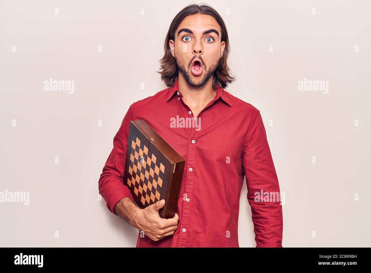 Young handsome man holding chess scared and amazed with open mouth for ...