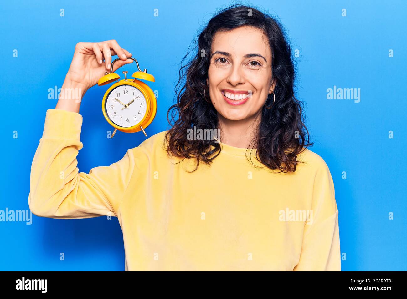 Young beautiful hispanic woman holding alarm clock looking positive and ...