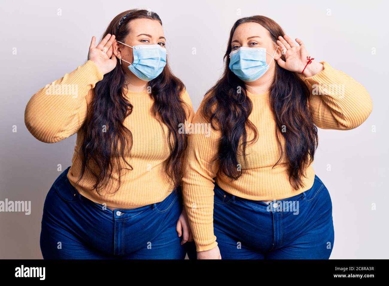 Young plus size twins wearing medical mask smiling with hand over ear ...