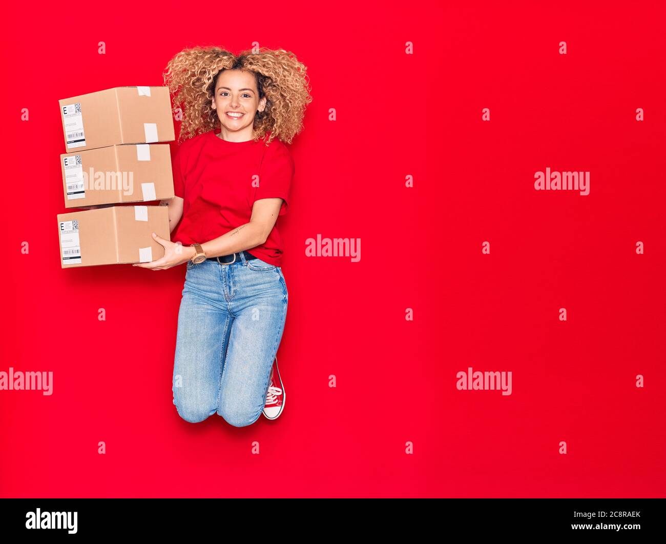 Young beautiful curly deliverywoman holding package smiling happy ...