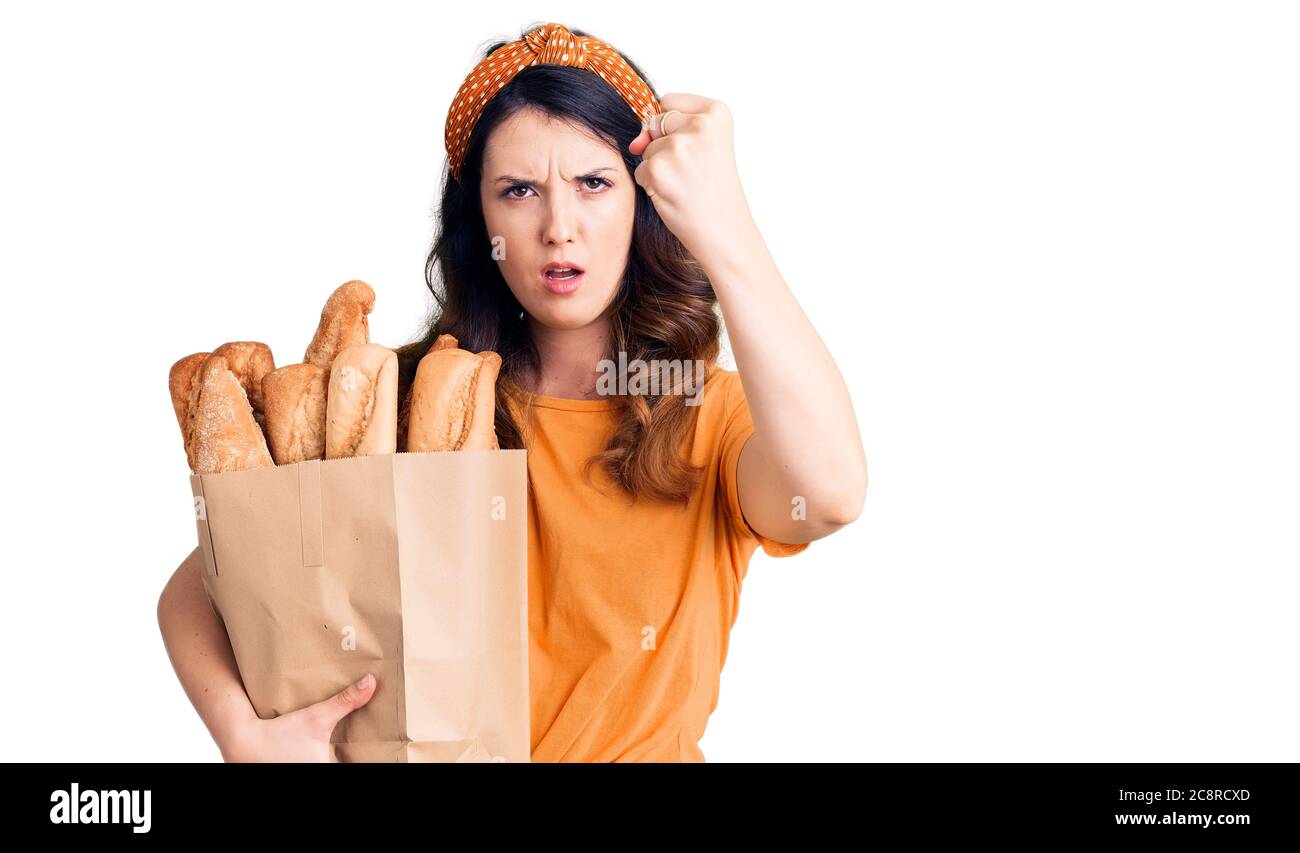 Beautiful young brunette woman holding paper bag with bread annoyed and ...
