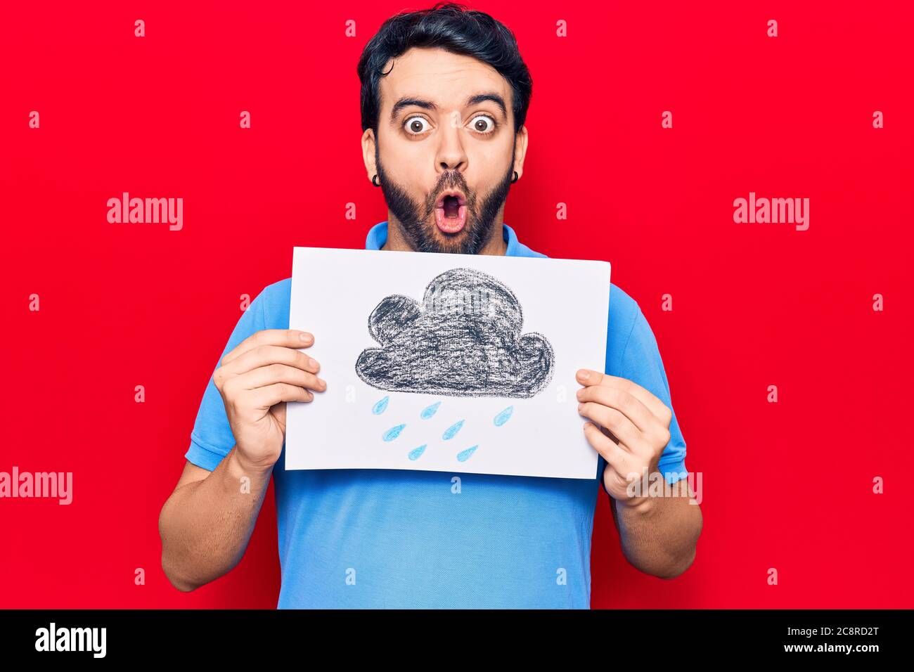 Young hispanic man holding rain draw scared and amazed with open mouth ...